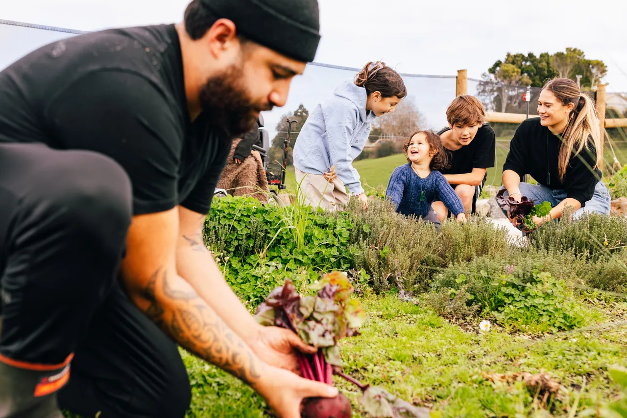 Man picks beetroot with family gardening in background
